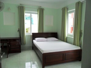 Quarto com uma cama de solteiro, escrivaninha e uma cadeira. Existem duas janelas com cortinas, e um ventilador fixado na parede.