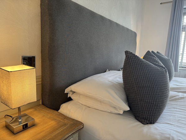 Cama bem arrumada com lençóis brancos e almofadas de textura escura, luminária de cabeceira e mesa de cabeceira de madeira.