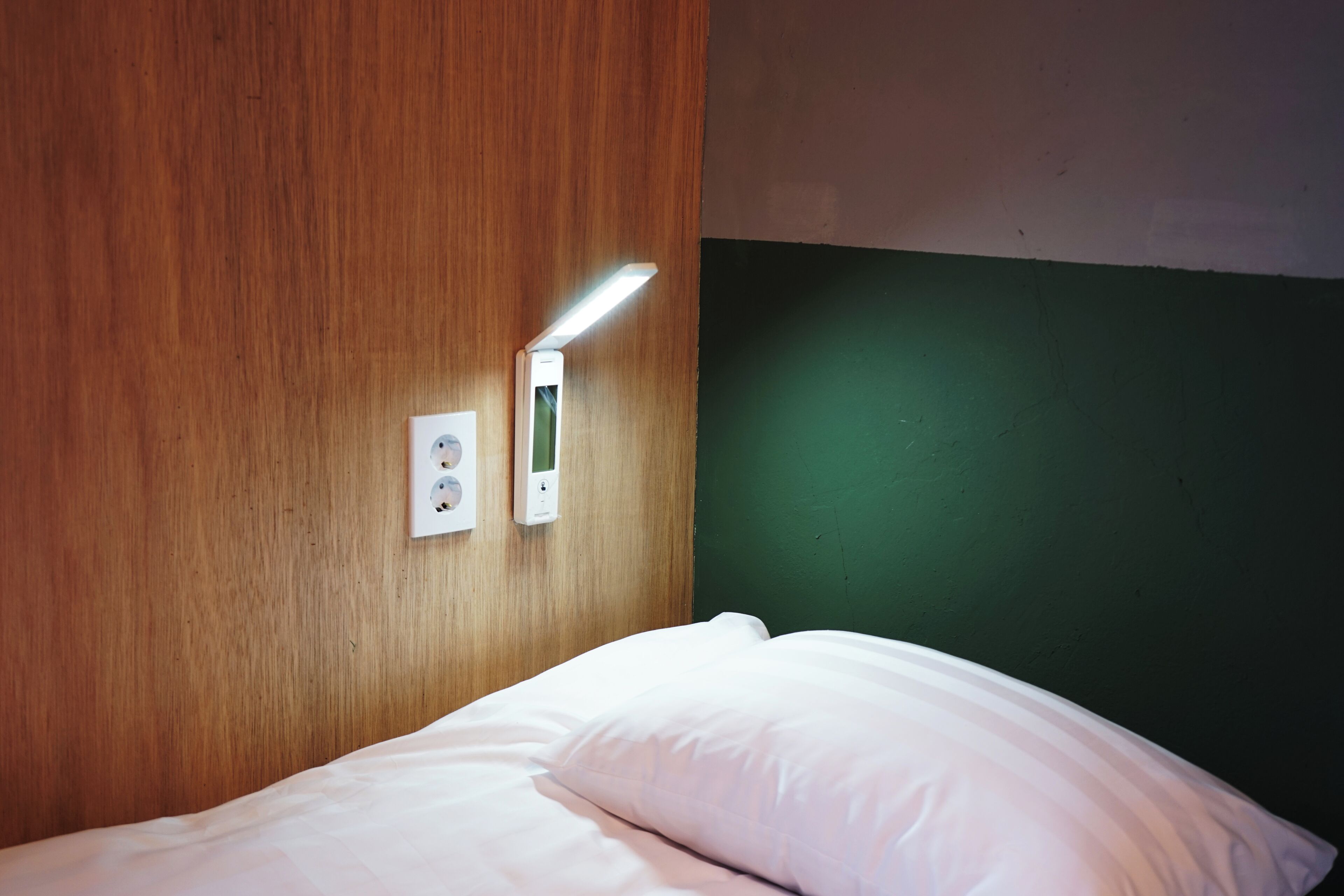 Quarto de hotel com cabeceira de madeira, cama branca e luminária fixada na parede.