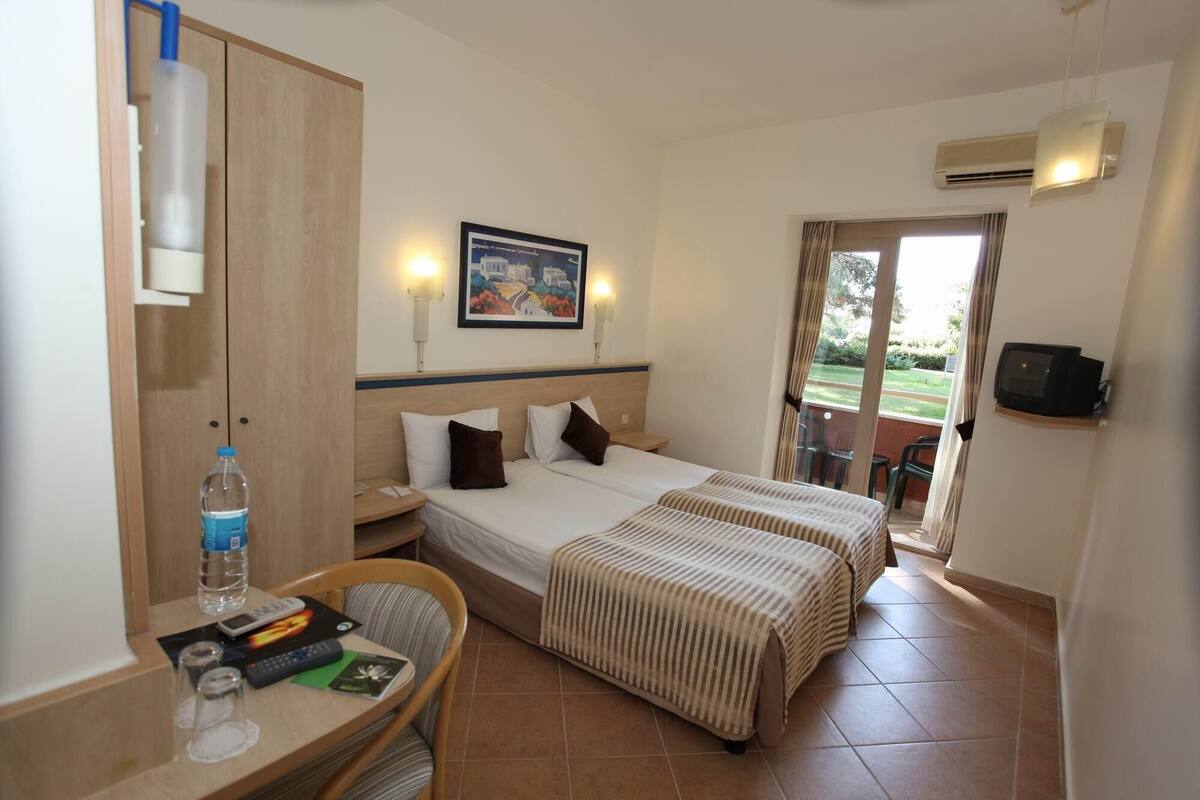 Quarto de hotel com duas camas, uma escrivaninha com cadeira, televisão e um quadro na parede.