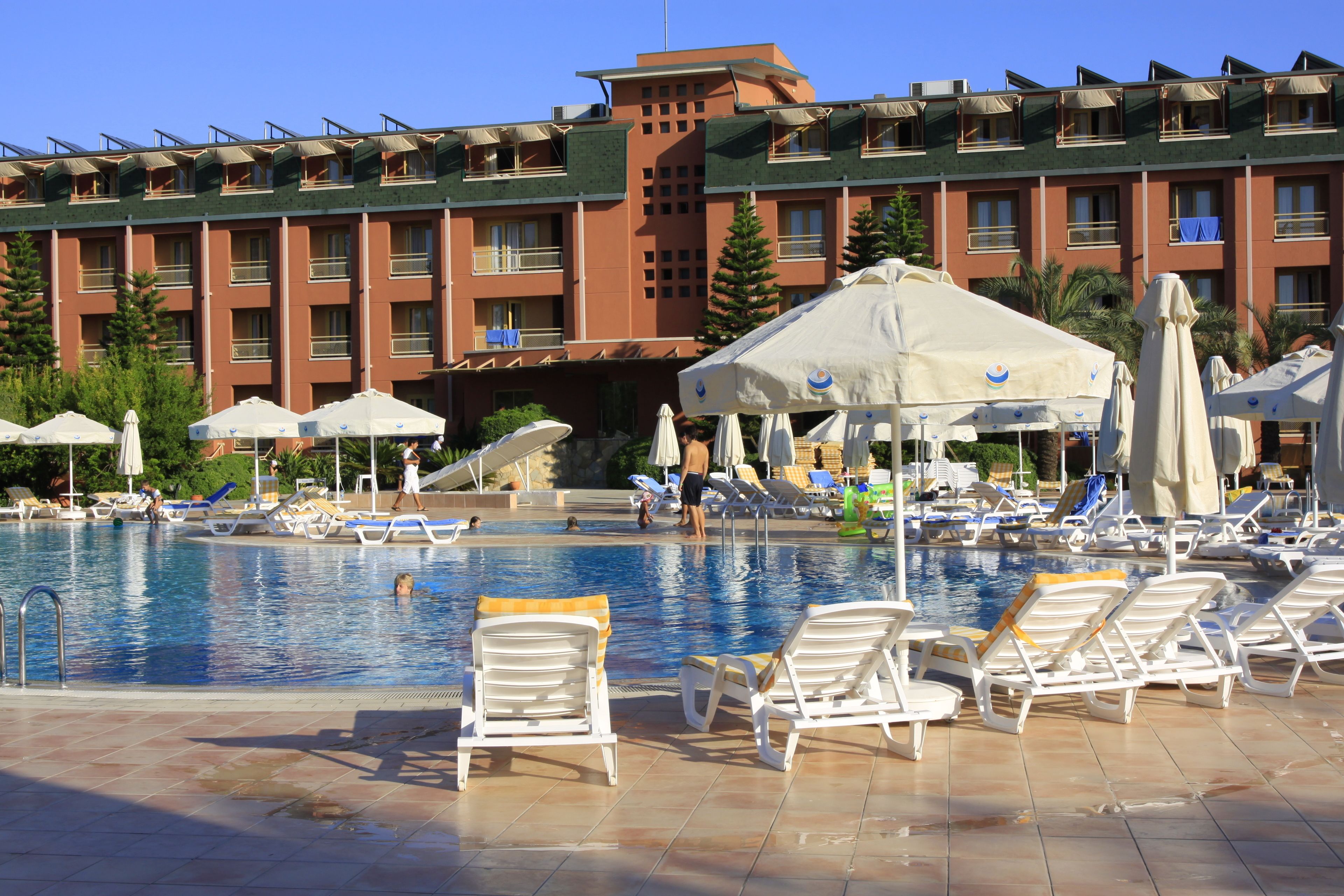 Uma área de piscina de hotel com espreguiçadeiras, guarda-sóis e um edifício grande ao fundo.