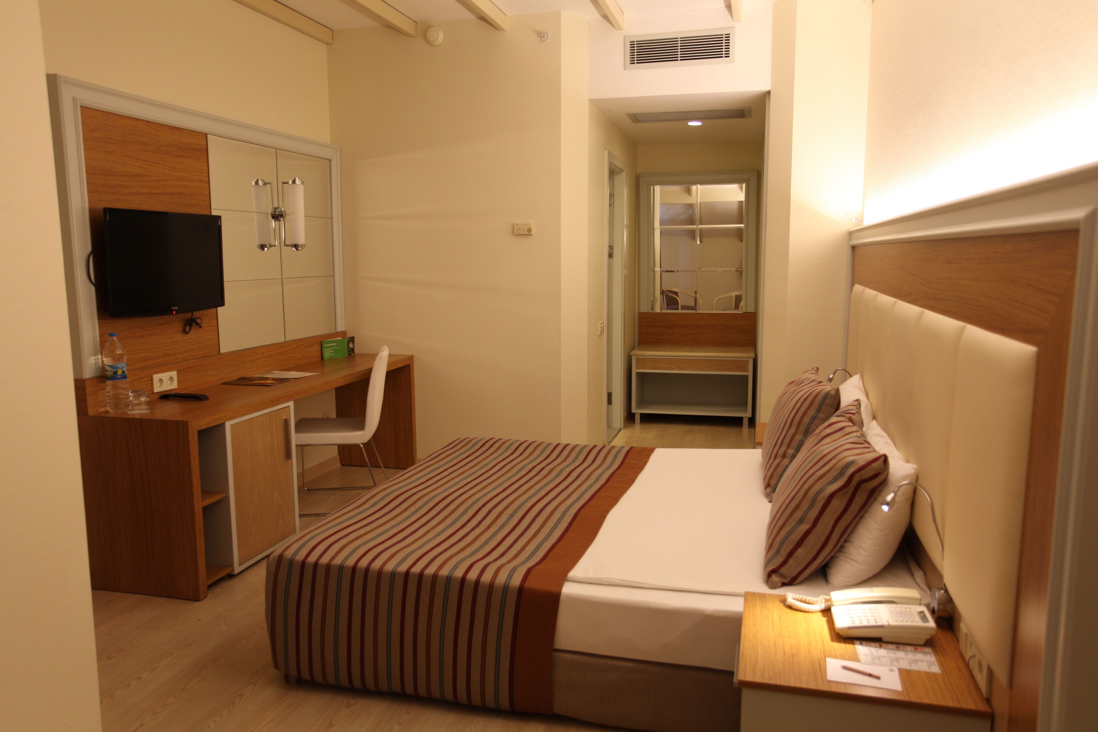 Quarto de hotel com cama, escrivaninha, cadeira, televisão e guarda-roupa.