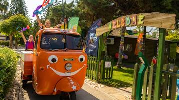 Um veículo laranja antigo com rosto sorridente e decorações de estrelas estacionado em frente a uma placa de madeira que diz “WELCOME”.