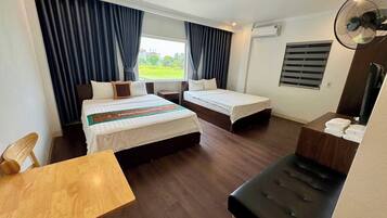 Quarto de hotel com duas camas, uma mesa de madeira, um banco, um ventilador e vista para área verde lá fora.