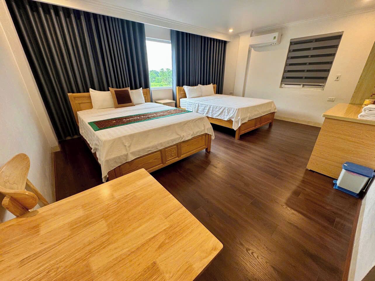 Quarto de hotel com duas camas, uma escrivaninha de madeira, uma cadeira, uma mesinha e uma janela com cortinas.