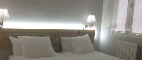 Uma cama de casal com roupa de cama branca e uma almofada escura. Há duas mesas de cabeceira com luminárias e uma janela com persianas à direita.