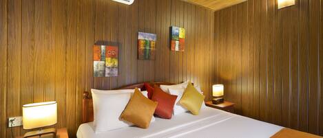 Quarto de hotel com parede revestida em madeira, uma cama com várias almofadas, mesa de cabeceira com abajur e ar condicionado.