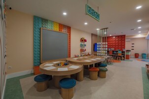 Uma sala de jogos infantil com mesas redondas, banquetas e brinquedos educativos.