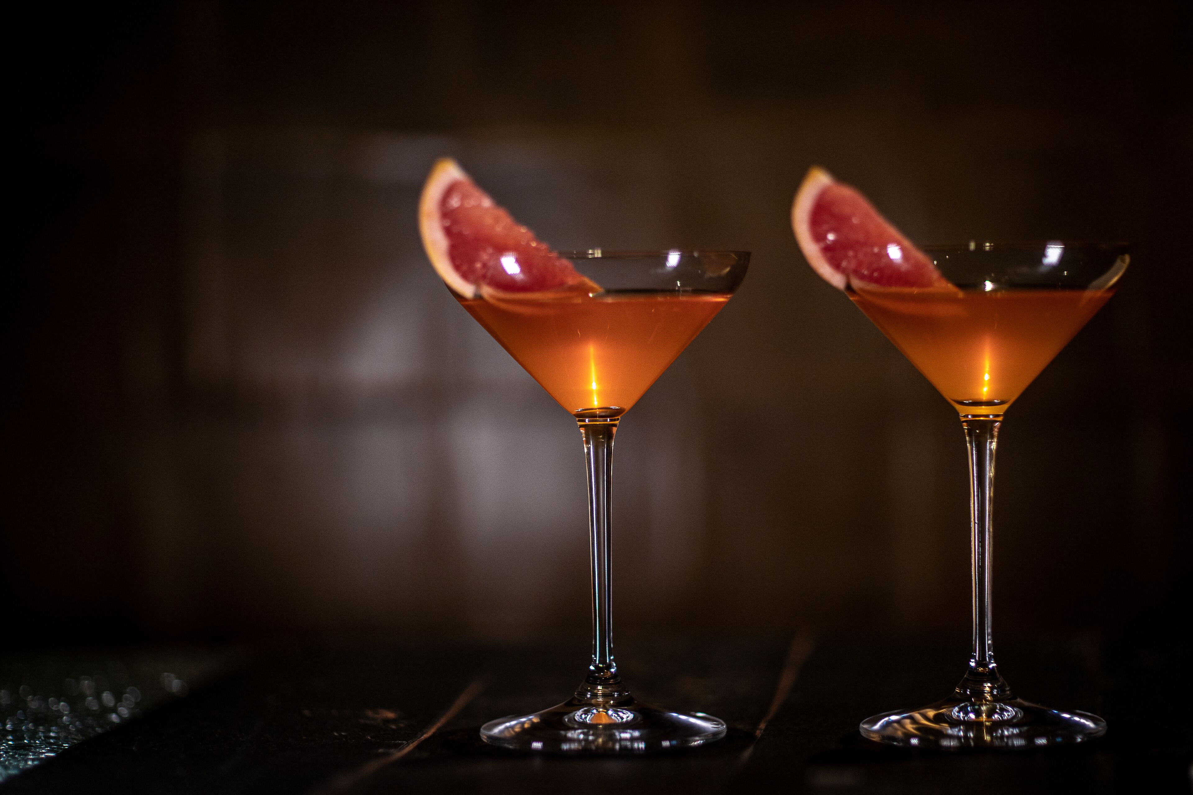 Dois copos de martini com líquido avermelhado-acastanhado, cada um decorado com uma fatia de laranja sanguínea, sobre uma superfície escura.