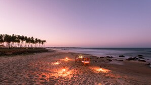 Cenário de praia com mesa e cadeiras montadas para refeições, rodeado por areia e palmeiras.
