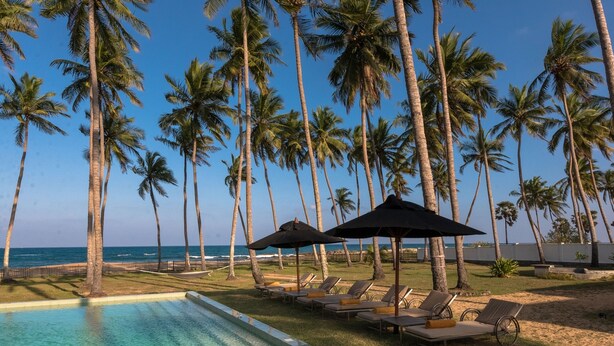 Área de piscina com água cristalina, rodeada por palmeiras e espreguiçadeiras sob guarda-sóis, com vista para o oceano.