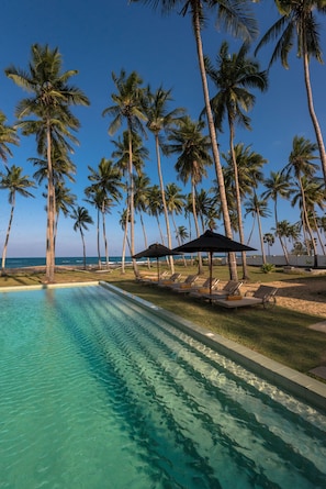 Área de piscina com água cristalina, rodeada por palmeiras e espreguiçadeiras sob guarda-sóis, com vista para o oceano.