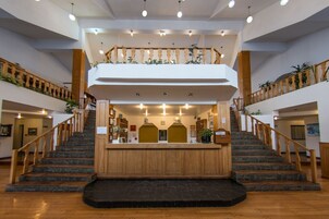 Um hall amplo com um balcão de recepção central, uma escadaria que leva a um mezanino e guarda-corpos de madeira.