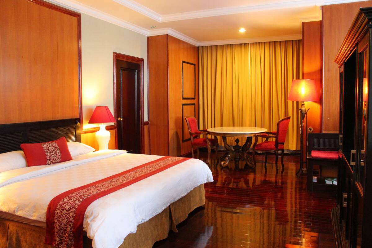 Quarto de hotel com uma cama grande, uma mesa de jantar com cadeiras e piso de madeira.