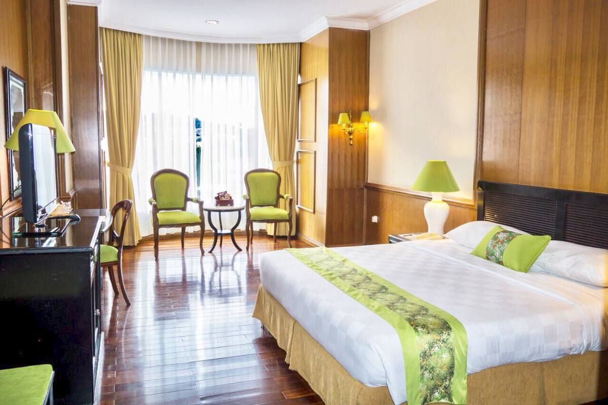 Quarto de hotel com uma cama grande, duas cadeiras, uma mesa pequena, uma escrivaninha com luminária e uma janela com cortinas.