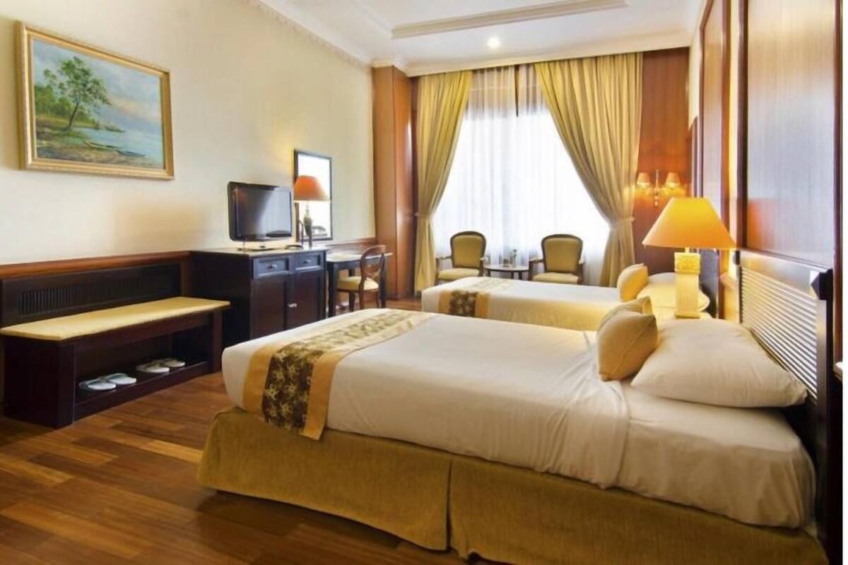 Quarto de hotel com duas camas, uma escrivaninha, uma cadeira, um banco e uma televisão.