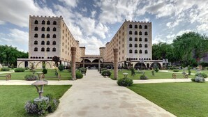 Um edifício grande e simétrico com um pátio central, apresentando um caminho pavimentado, áreas de estar e elementos decorativos como estátuas e vasos de plantas.