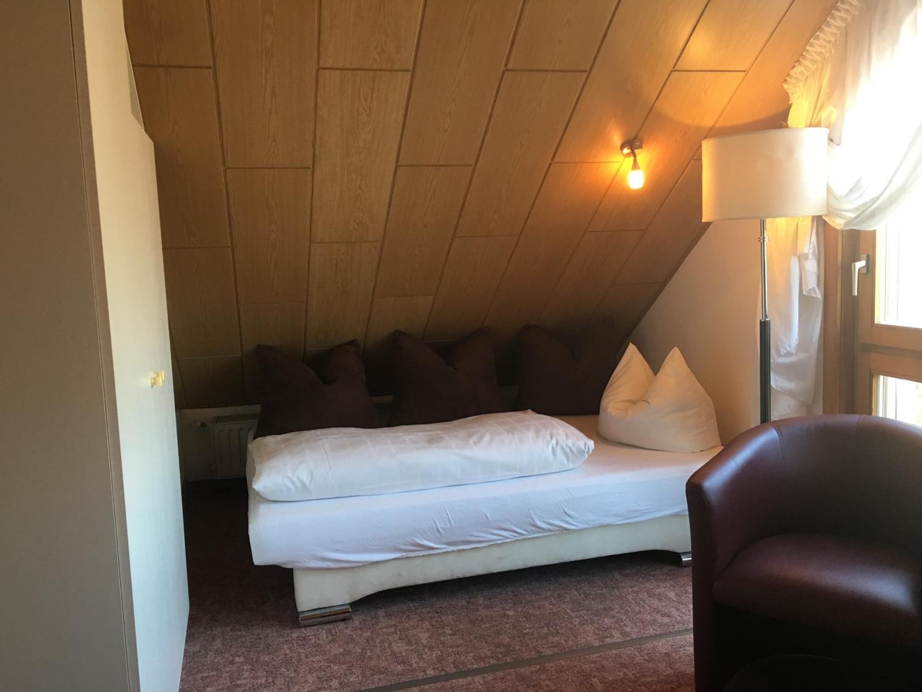 Quarto com cama, almofadas, uma cadeira, abajur e teto de madeira.