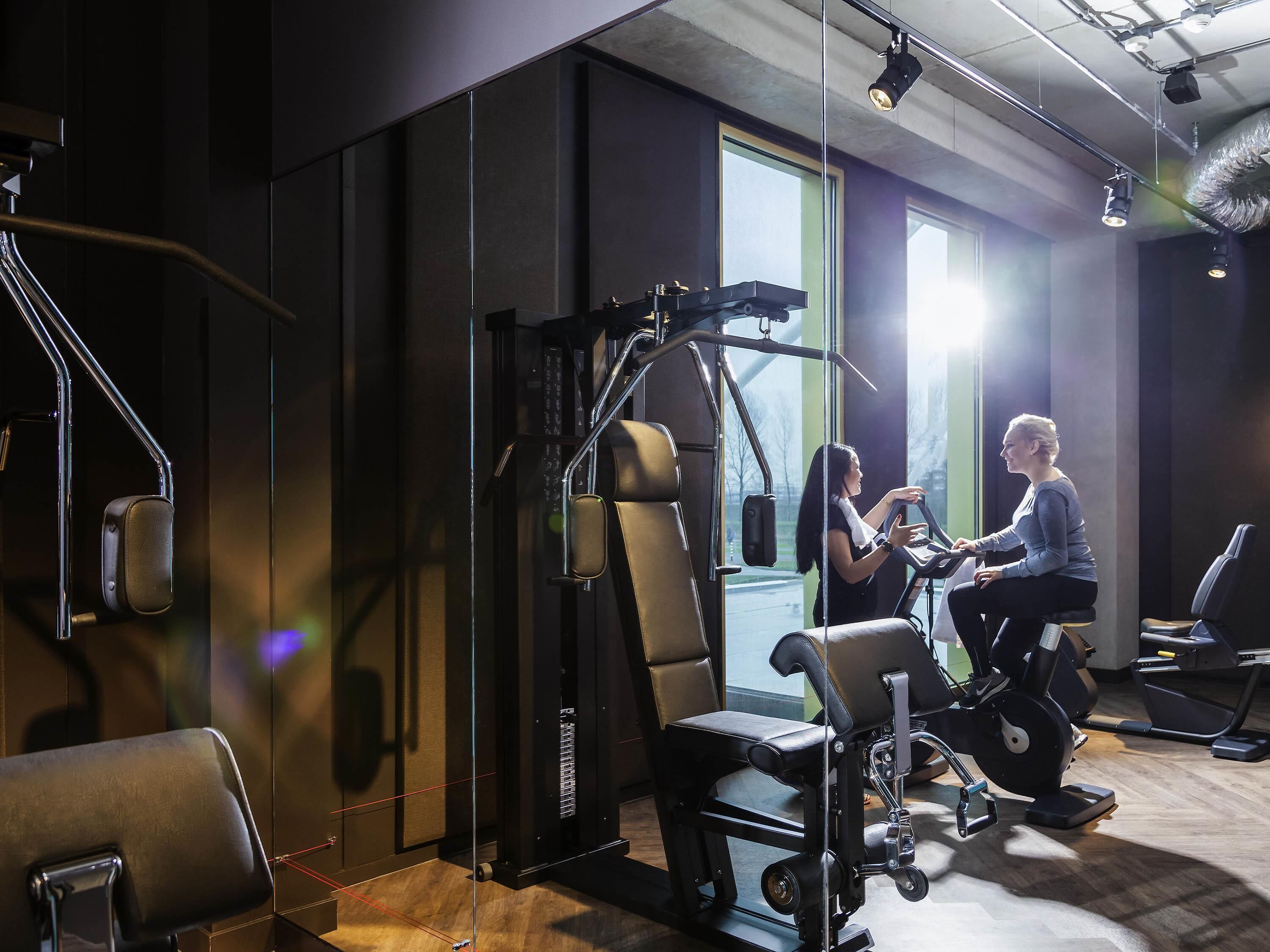 Uma academia com equipamentos de ginástica, esteira e janela com luz natural.