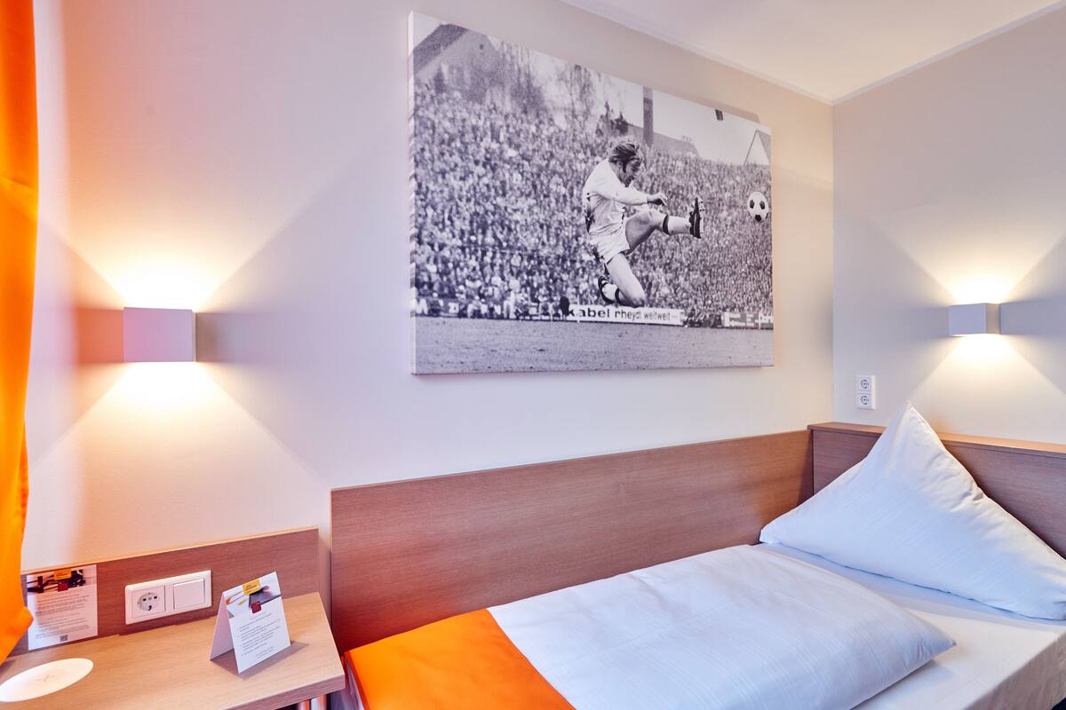 Quarto de hotel com cama, mesa de cabeceira, luminária, um pôster na parede e janela com cortinas.