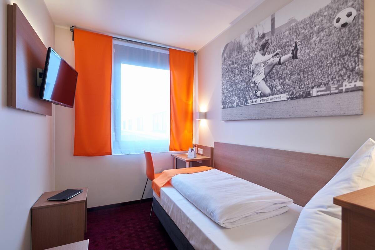 Quarto de hotel com cama, escrivaninha, cadeira, televisão fixada na parede e janela com cortinas.
