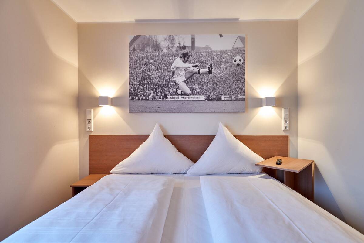 Quarto de hotel com uma cama, duas almofadas, mesa de cabeceira com controle remoto e uma foto emoldurada na parede.