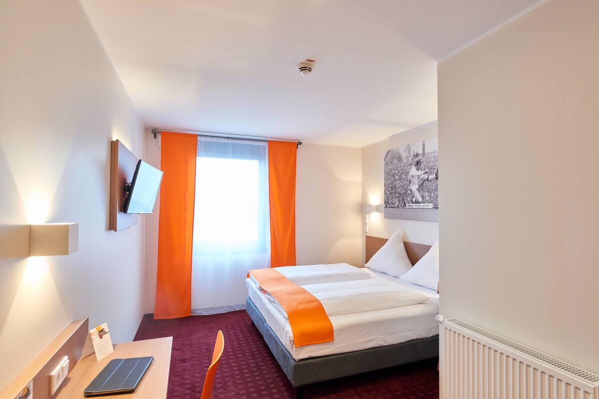 Quarto de hotel com duas camas, uma secretária com cadeira, televisão de tela plana e uma janela com cortinas laranja.