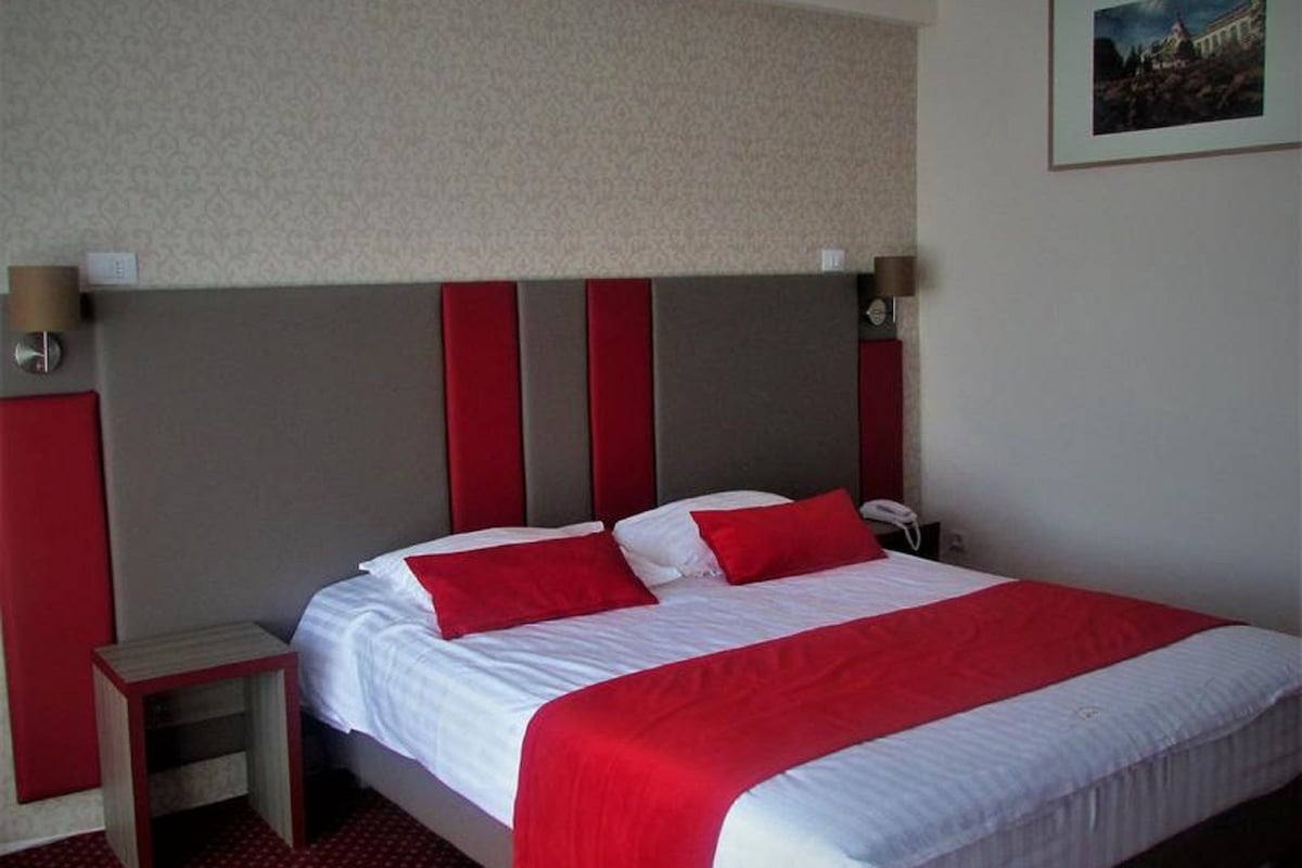 Quarto de hotel com uma cama grande, roupa de cama vermelha e branca, mesa de cabeceira e um quadro na parede.