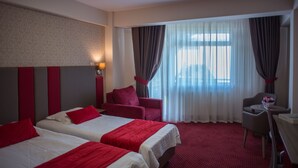Quarto de hotel com duas camas, uma escrivaninha, uma cadeira e uma janela grande com cortinas.