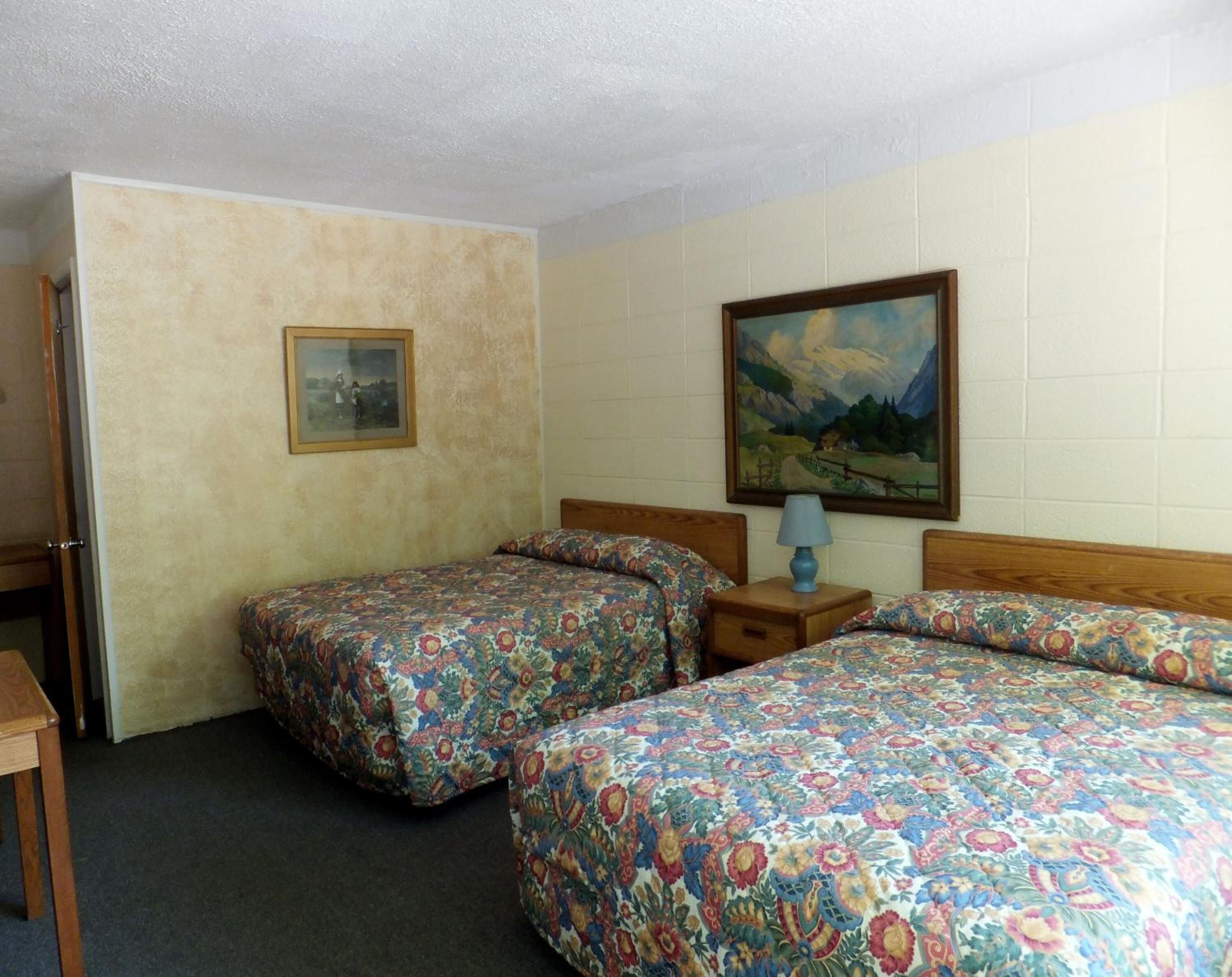 Quarto com duas camas de solteiro, uma mesa de cabeceira de madeira com abajur, um quadro de paisagem na parede e uma porta que dá para outro quarto.