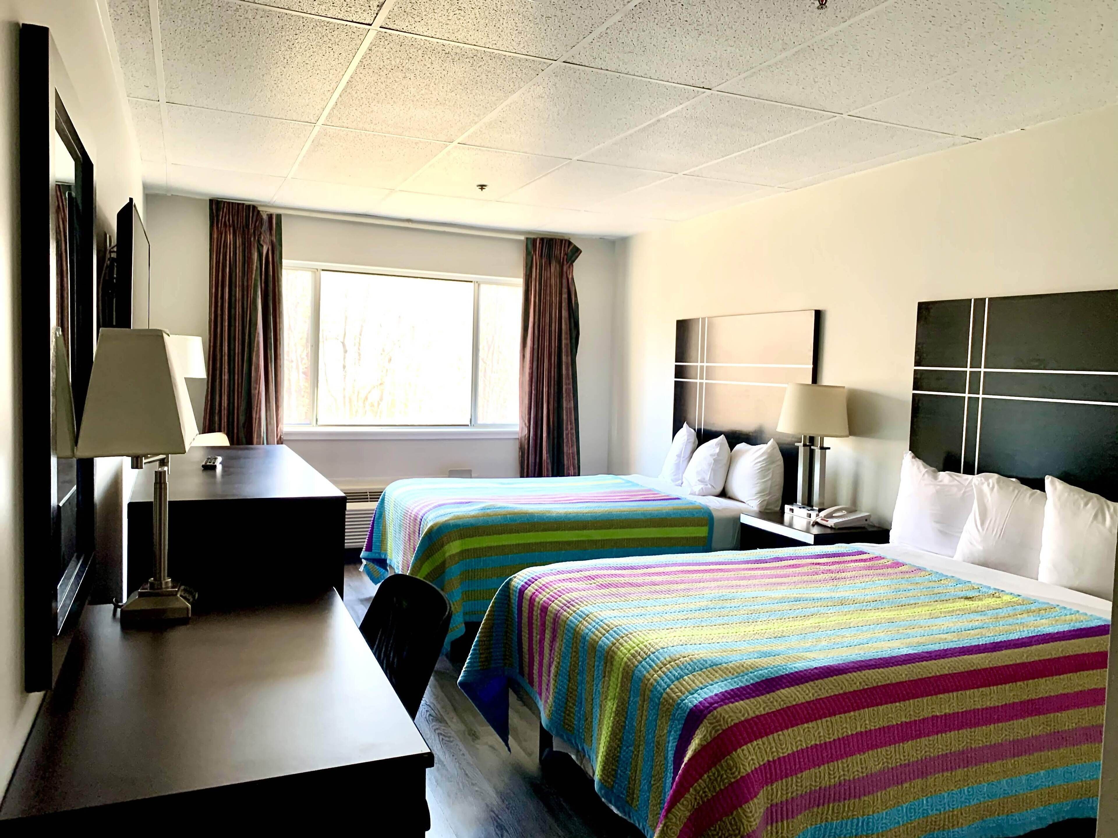 Quarto de hotel com duas camas, uma mesa, uma cadeira, mesa de cabeceira e janela com cortinas.