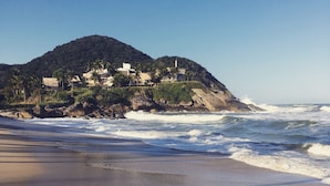 Uma praia com costa rochosa, ondas quebrando na areia, e uma colina com edifícios e palmeiras ao fundo.