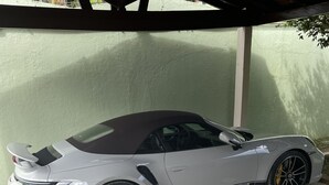Um Porsche conversível prateado estacionado em área coberta com piso de concreto e parede verde ao fundo.
