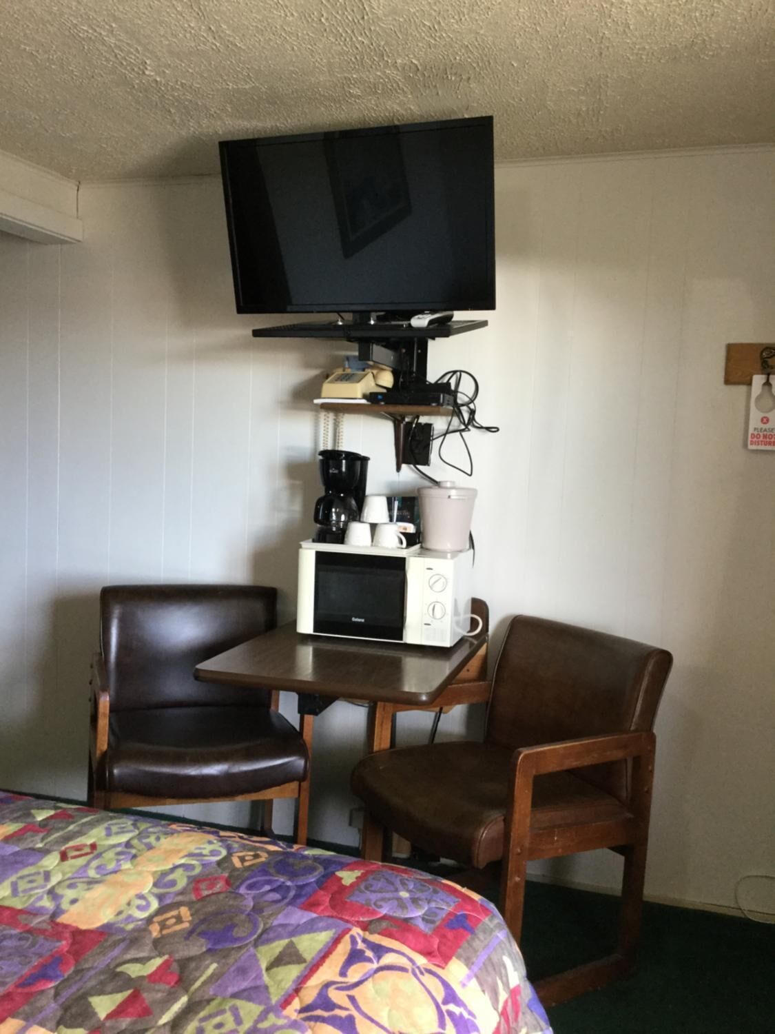 Quarto de hotel com cama, duas cadeiras, uma mesinha com micro-ondas, máquina de café e televisão fixada na parede.