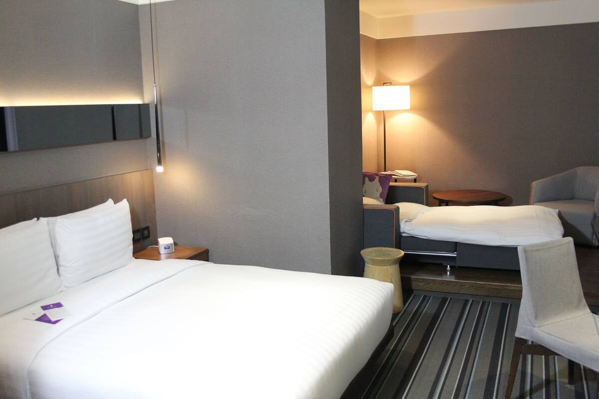 Quarto de hotel com uma cama grande, uma mesa de cabeceira com abajur, uma cadeira e uma mesinha com telefone.