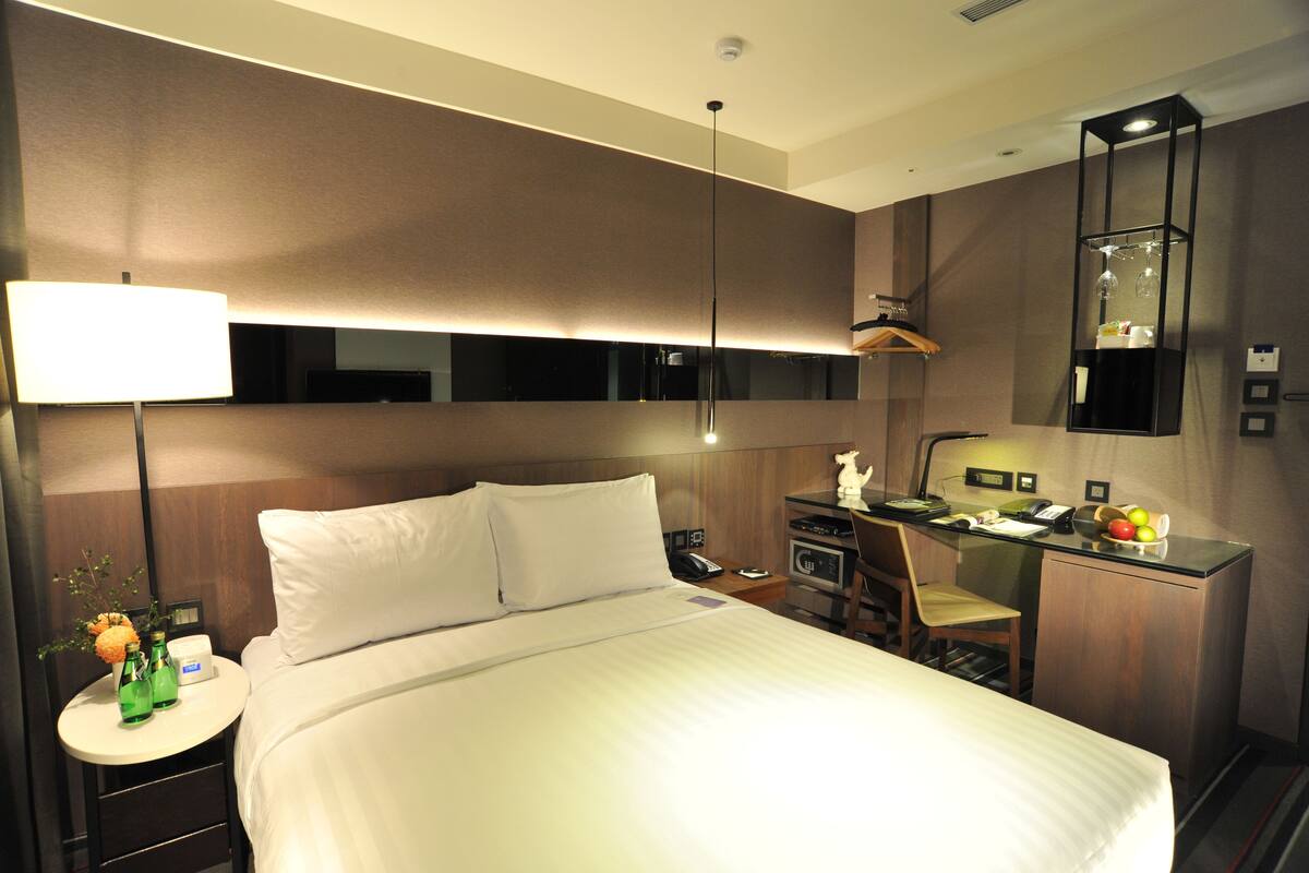Quarto de hotel com uma cama, uma escrivaninha, uma cadeira, um abajur, um espelho e uma mesinha com um vaso e garrafas.