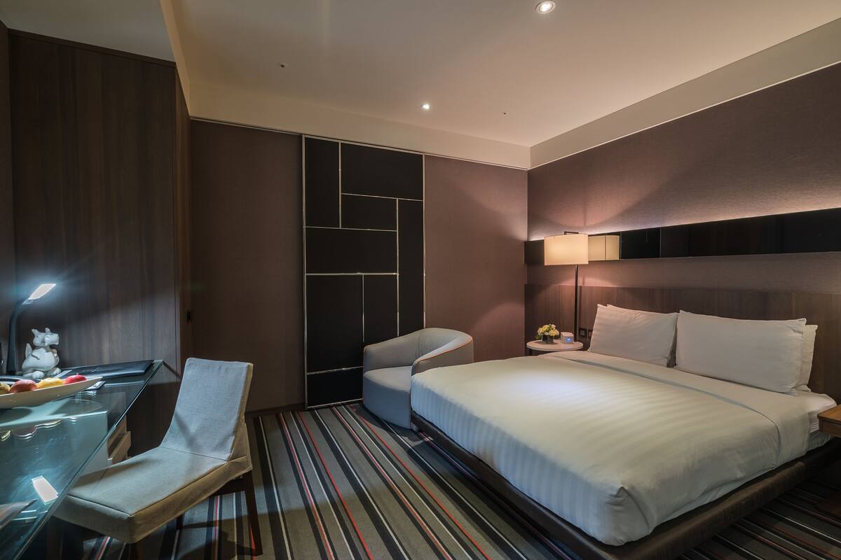 Quarto de hotel moderno com uma cama grande, uma escrivaninha de vidro, uma cadeira e uma mesinha com frutas.
