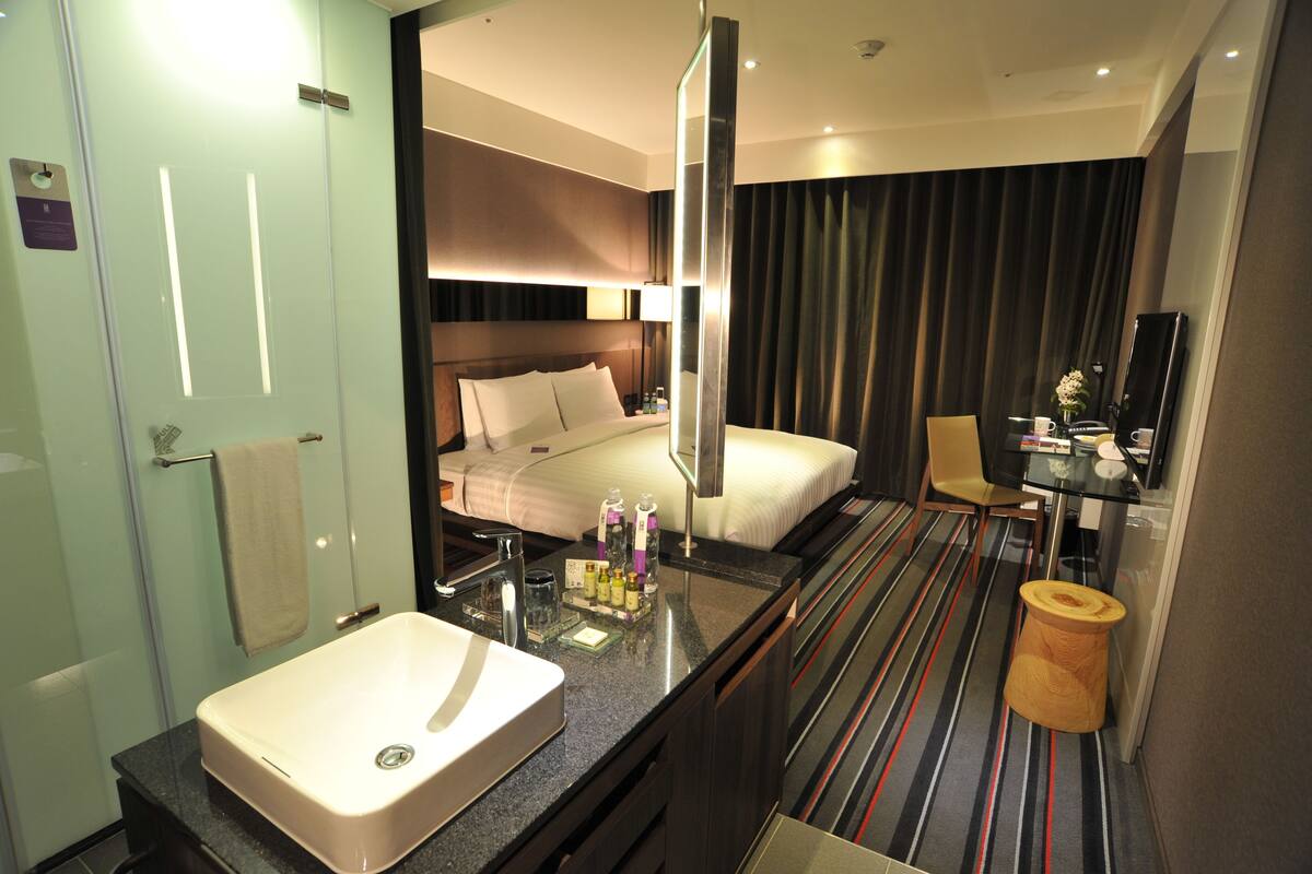 Quarto de hotel com cama, banheiro com pia, espelho, cadeira e mesa pequena.
