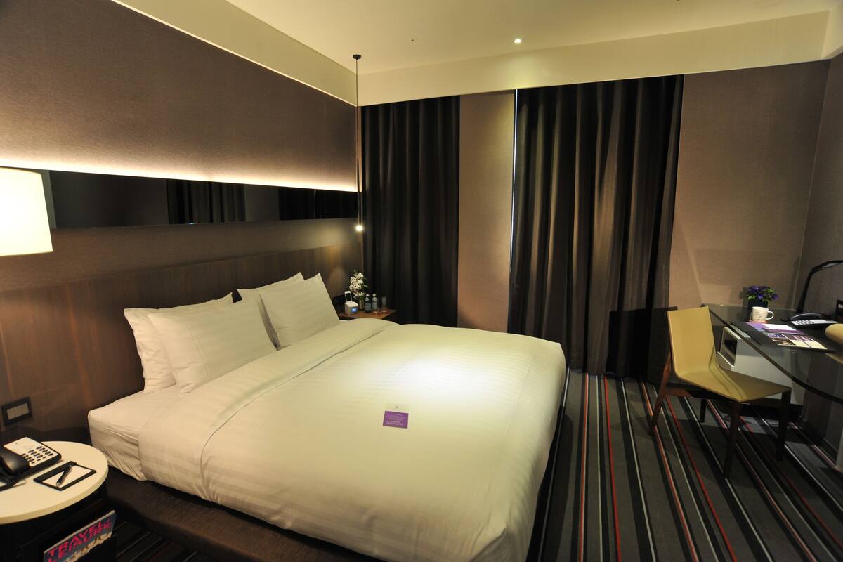 Quarto de hotel com uma cama grande, uma escrivaninha com cadeira, telefone e mesa de cabeceira.