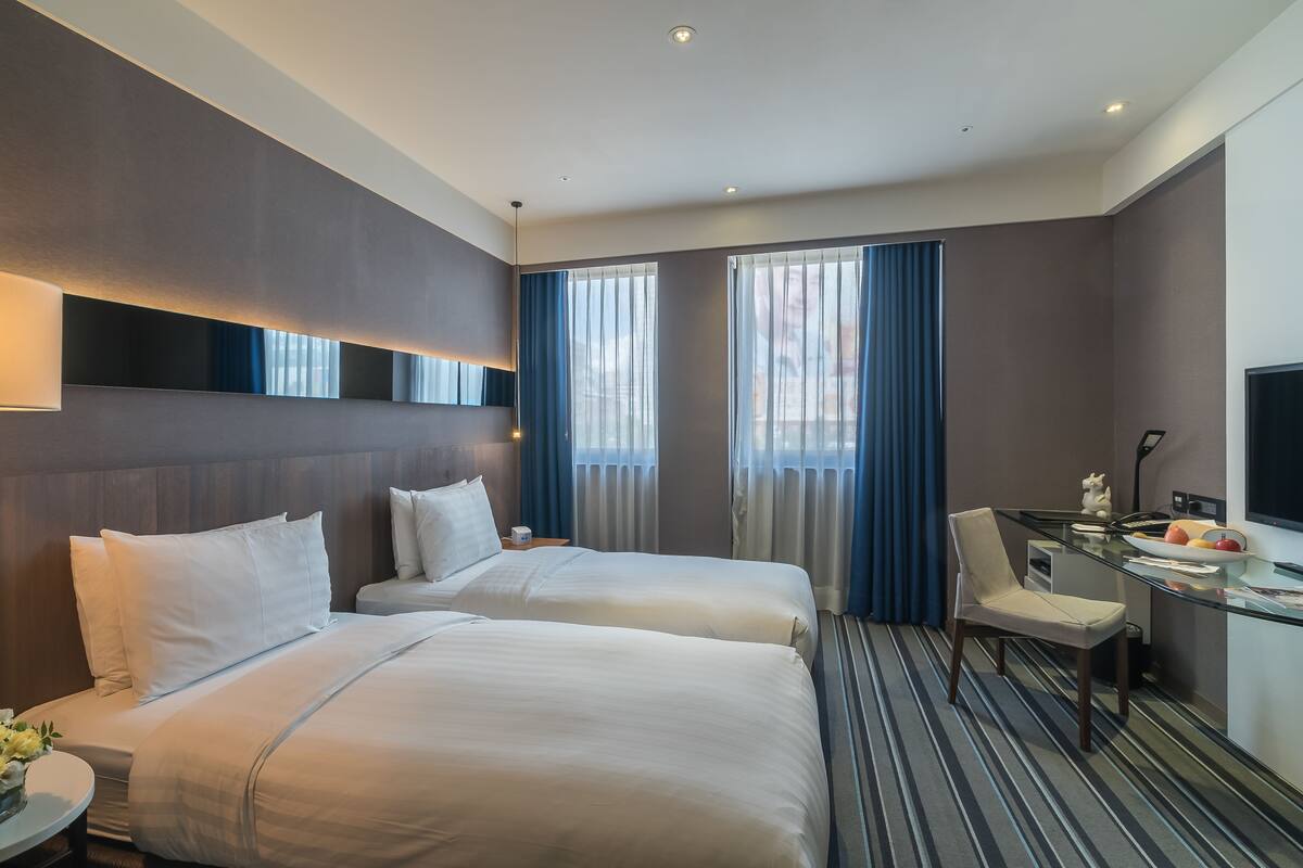 Quarto de hotel com duas camas, uma escrivaninha, uma cadeira, uma televisão e uma janela com cortinas.