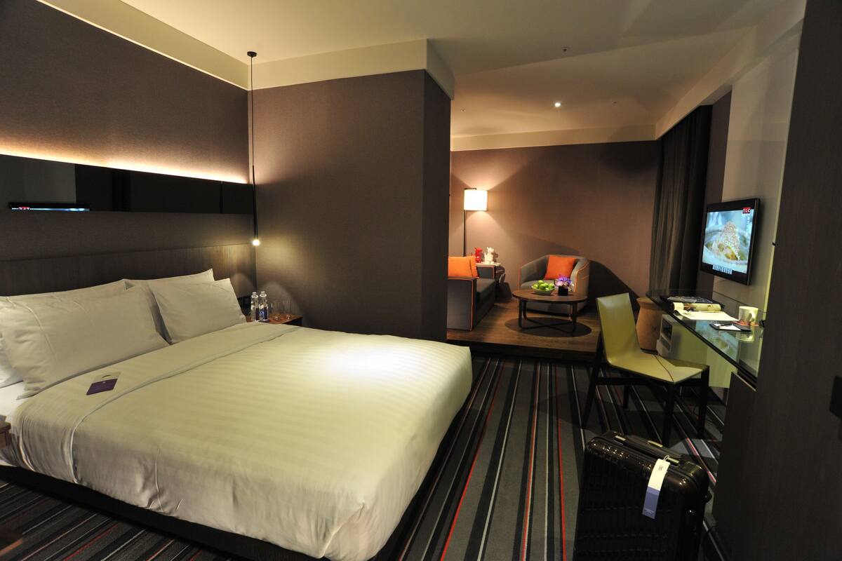 Quarto de hotel com uma cama grande, uma escrivaninha com cadeira, uma televisão e uma mesinha de cabeceira com abajur.