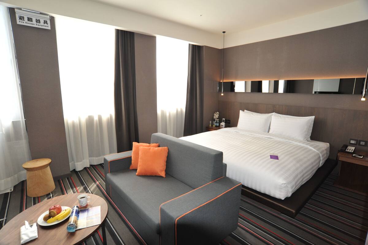 Quarto de hotel com cama, sofá, uma pequena mesa com uma travessa de fruta, uma xícara e uma garrafa.