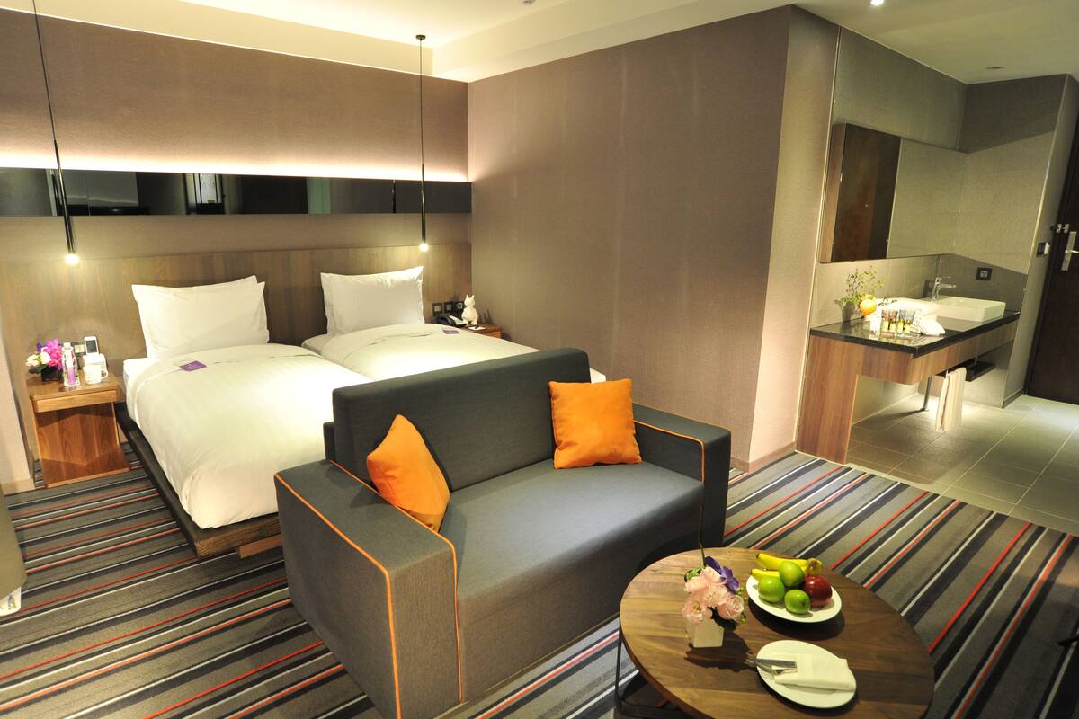 Quarto de hotel com cama, sofá, mesa de jantar com frutas e banheiro com pia e espelho.