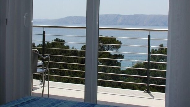 Quarto com cama, cadeira e varanda com vista para o mar e montanhas.