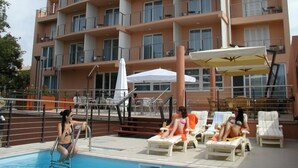 Área da piscina com espreguiçadeiras e guarda-sóis, e um edifício com várias varandas ao fundo.