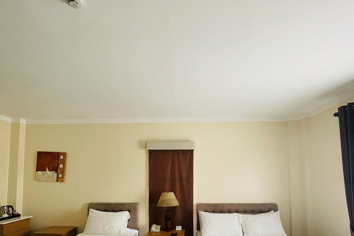 Quarto de hotel com duas camas de solteiro, uma mesinha de cabeceira com abajur, telefone e uma pequena mesa com uma xícara e um livro.