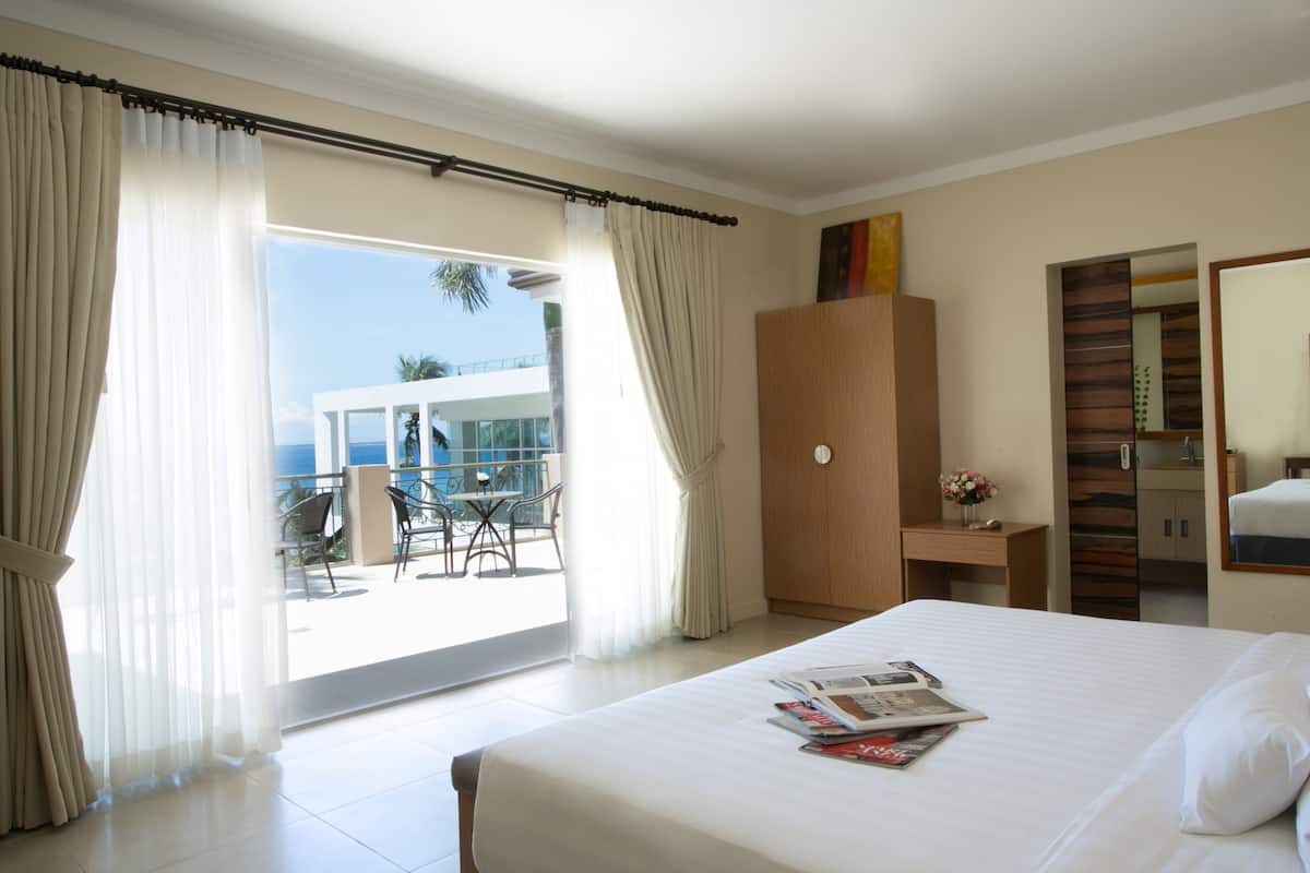 Quarto de hotel com cama, uma pequena escrivaninha, espelho e vista para o oceano.