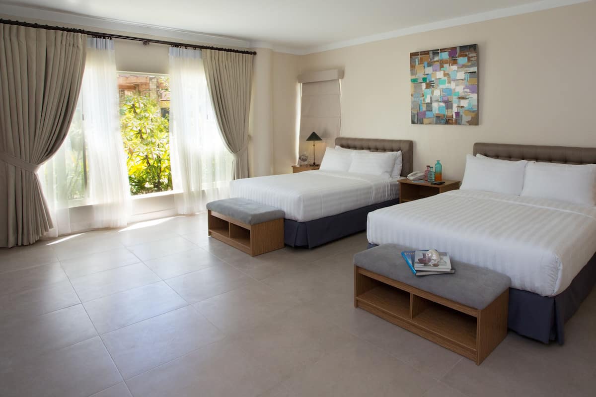 Quarto de hotel com duas camas, um banco, uma pintura e vista para o verde lá fora.