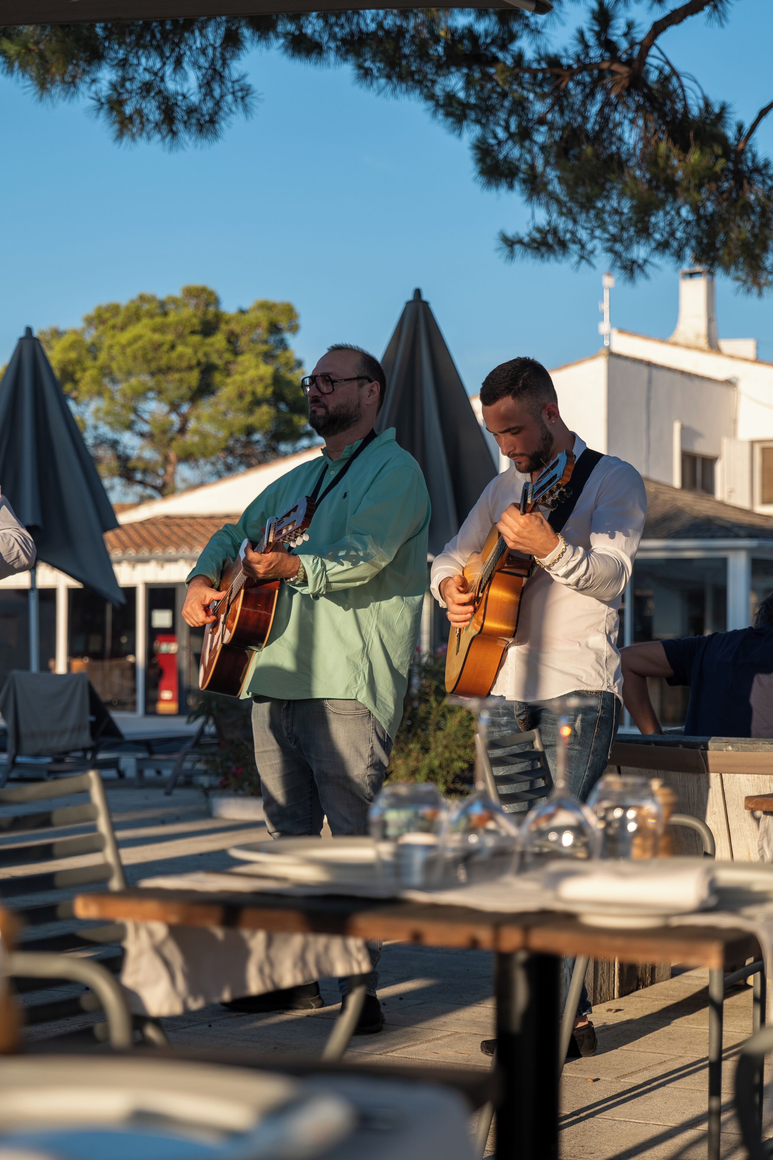 Dois homens tocando violões em uma mesa ao ar livre, com cadeiras e guarda-sóis ao fundo.