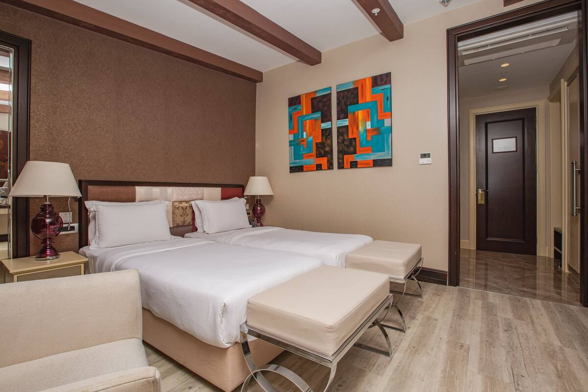 Quarto de hotel com uma cama grande, duas mesas de cabeceira com luminárias, um banco e uma porta que dá para outro quarto.